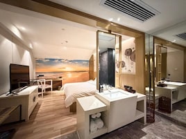 Quarto de hotel moderno com uma cama grande, uma mesa de trabalho, um banheiro com um espelho amplo e vista para o pôr do sol.