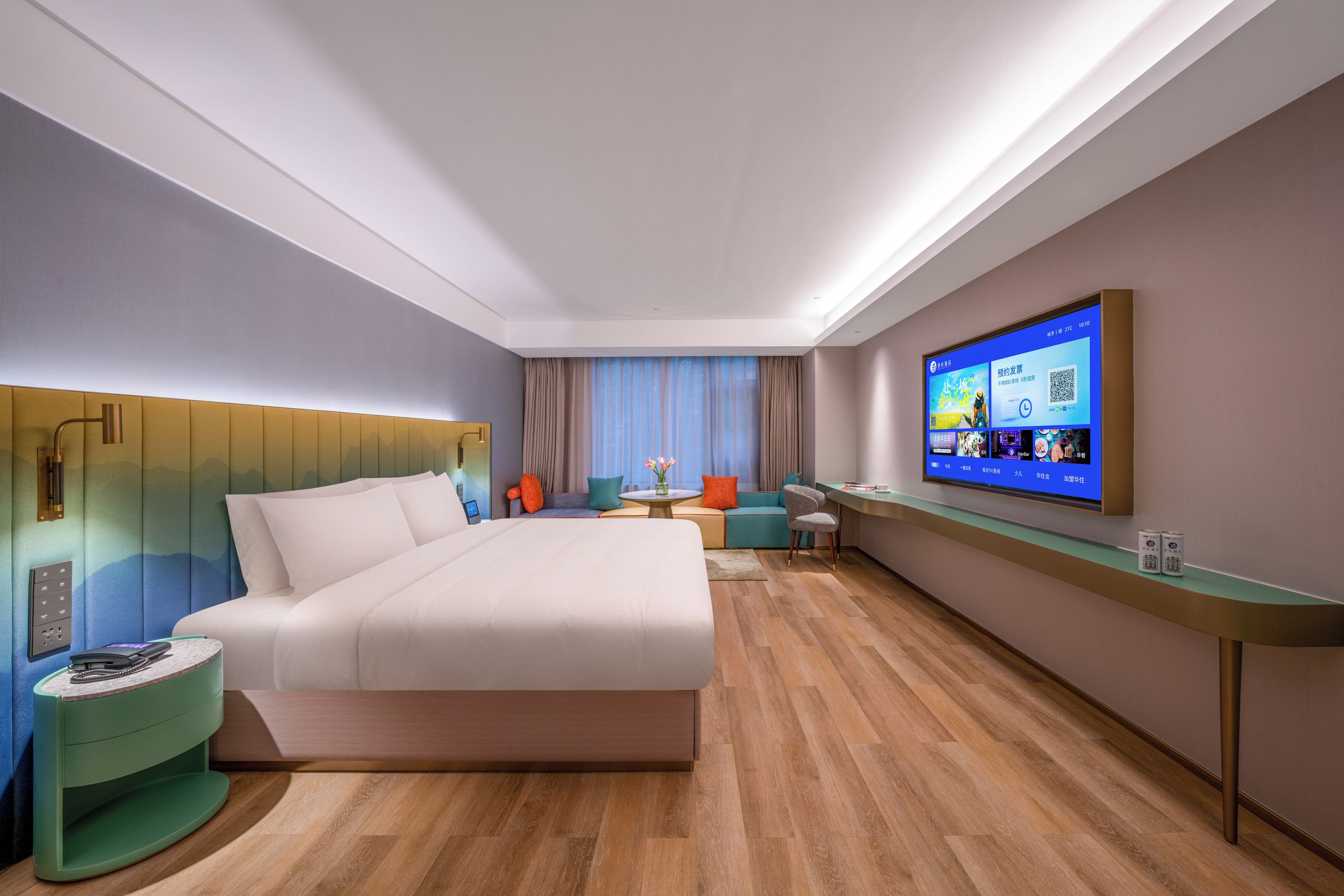Quarto de hotel moderno com uma cama grande, uma escrivaninha com televisão de tela plana e um espaço de estar com sofá e poltronas.