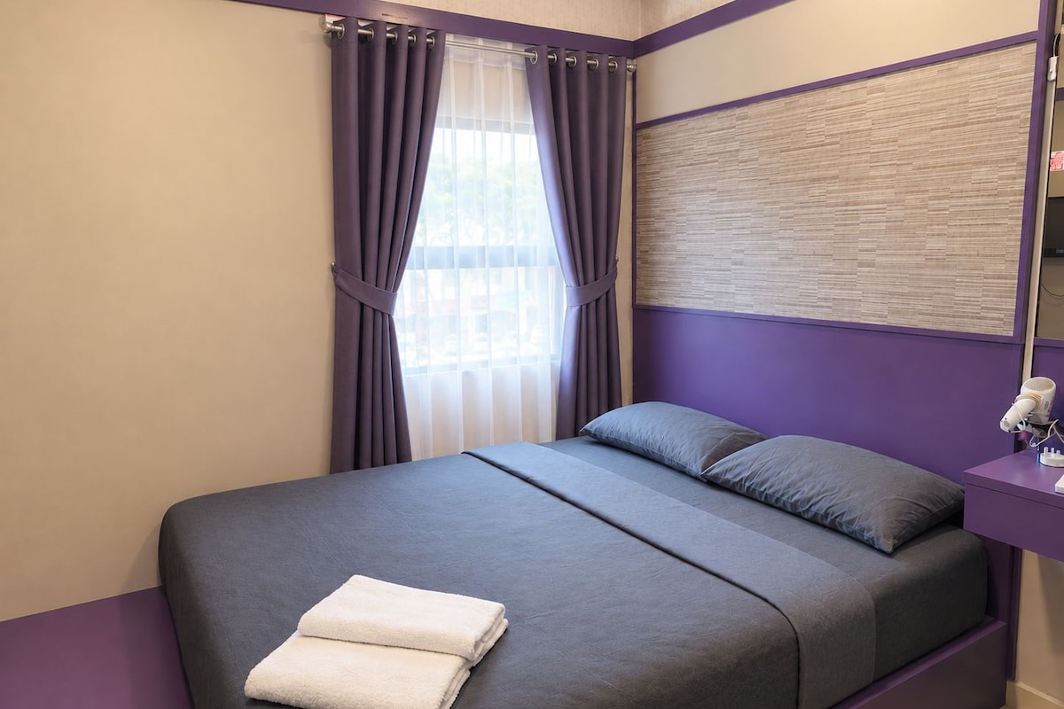 Quarto com cama, almofadas, janela com cortinas, mesa de cabeceira com abajur e toalha sobre a cama.