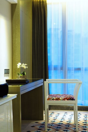 Quarto de hotel com uma janela grande, uma cadeira branca com almofada colorida, uma mesinha com um vaso de flores e um chão estampado.