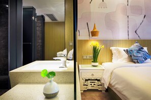 Quarto de hotel com banheiro, cama com travesseiro, mesa de cabeceira, vaso com flores e um quadro na parede.