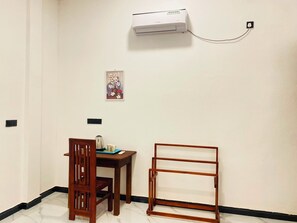 Quarto com uma cadeira de madeira, uma pequena mesa e um suporte de metal encostado em uma parede lisa.