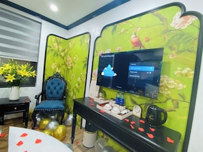 Quarto com televisão, uma cadeira azul, um vaso com flores amarelas e uma mesa com diversos objetos.