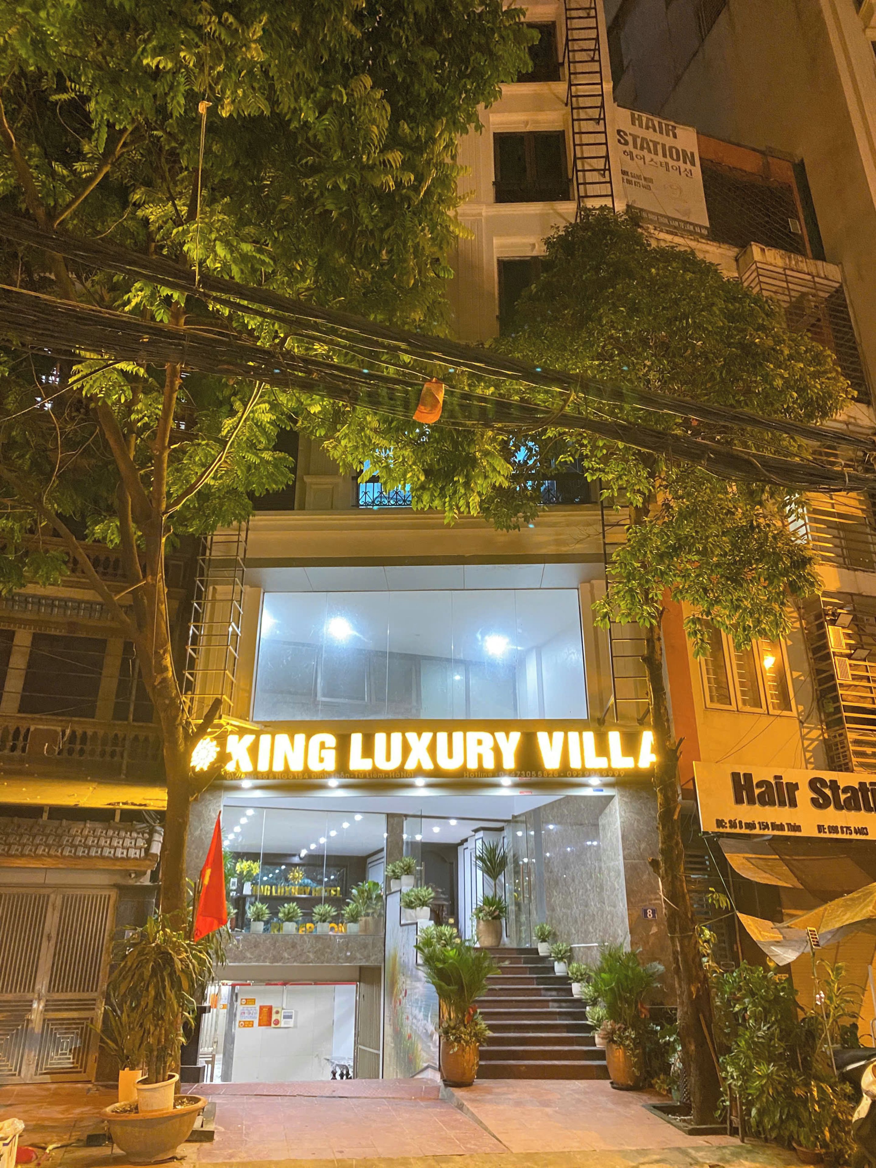 Um edifício com placas indicando 'KING LUXURY VILLA' e 'HAIR STATION'. Há plantas em vasos e uma escadaria que leva à entrada.