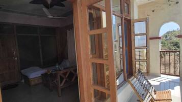 Quarto com porta de madeira, ventilador de teto, cama, mesa com cadeira e vista para o exterior.