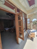Quarto com porta de madeira, ventilador de teto, cama, mesa com cadeira e vista para o exterior.