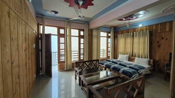 Quarto com cama, mesa e ventilador de teto. Possui cortinas nas janelas e uma porta de varanda.