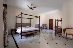 Uma cama de dossel com ventoinha no teto, uma cadeira, uma mesa e uma porta em quarto de hotel com piso de azulejos.
