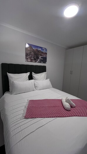 Uma cama bem arrumada com lençóis e travesseiros brancos, um cobertor xadrez vermelho e uma toalha enrolada sobre a cama.