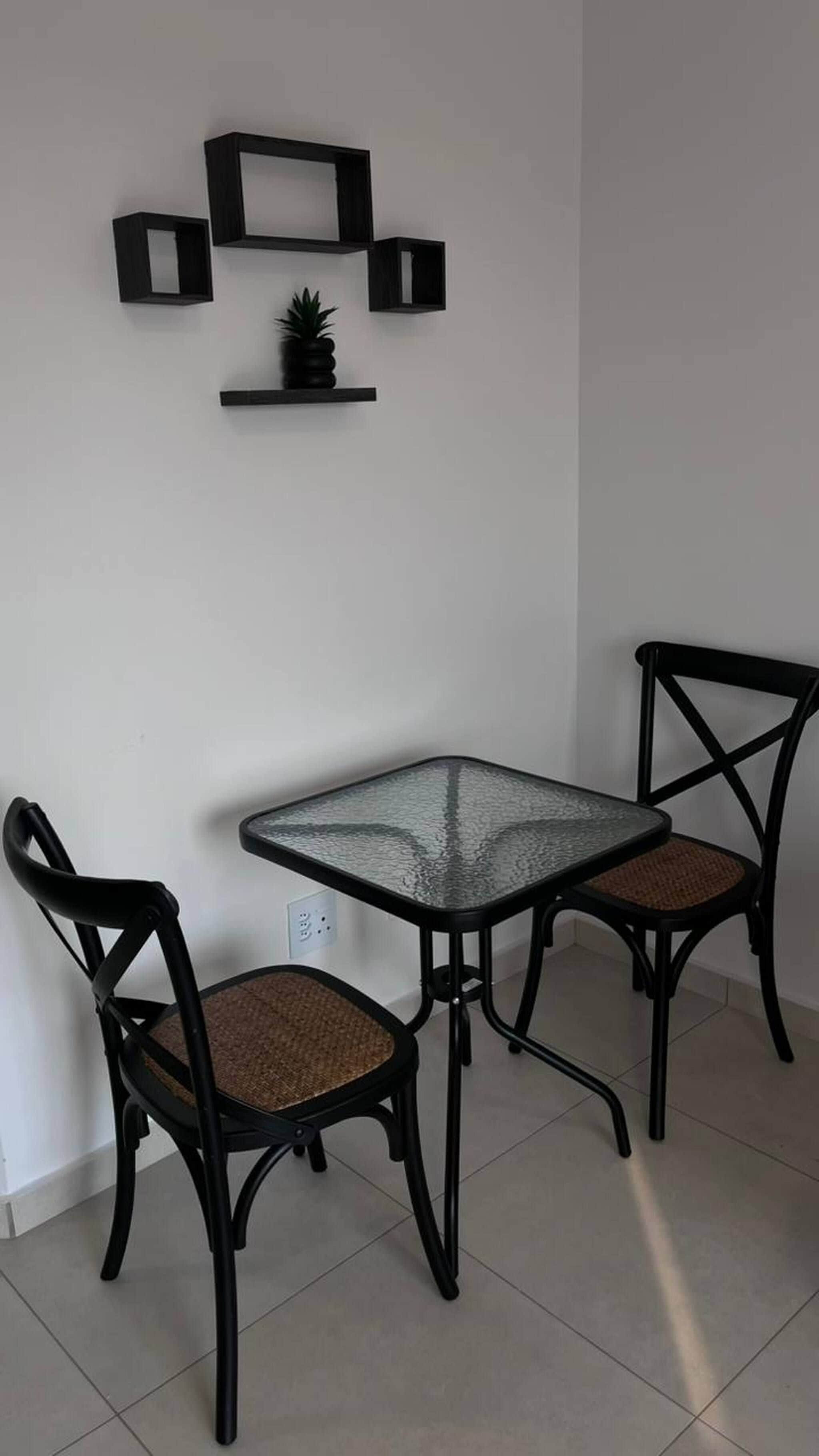 Quarto de canto com mesa de vidro e duas cadeiras, uma prateleira pequena com uma planta e uma tomada na parede.