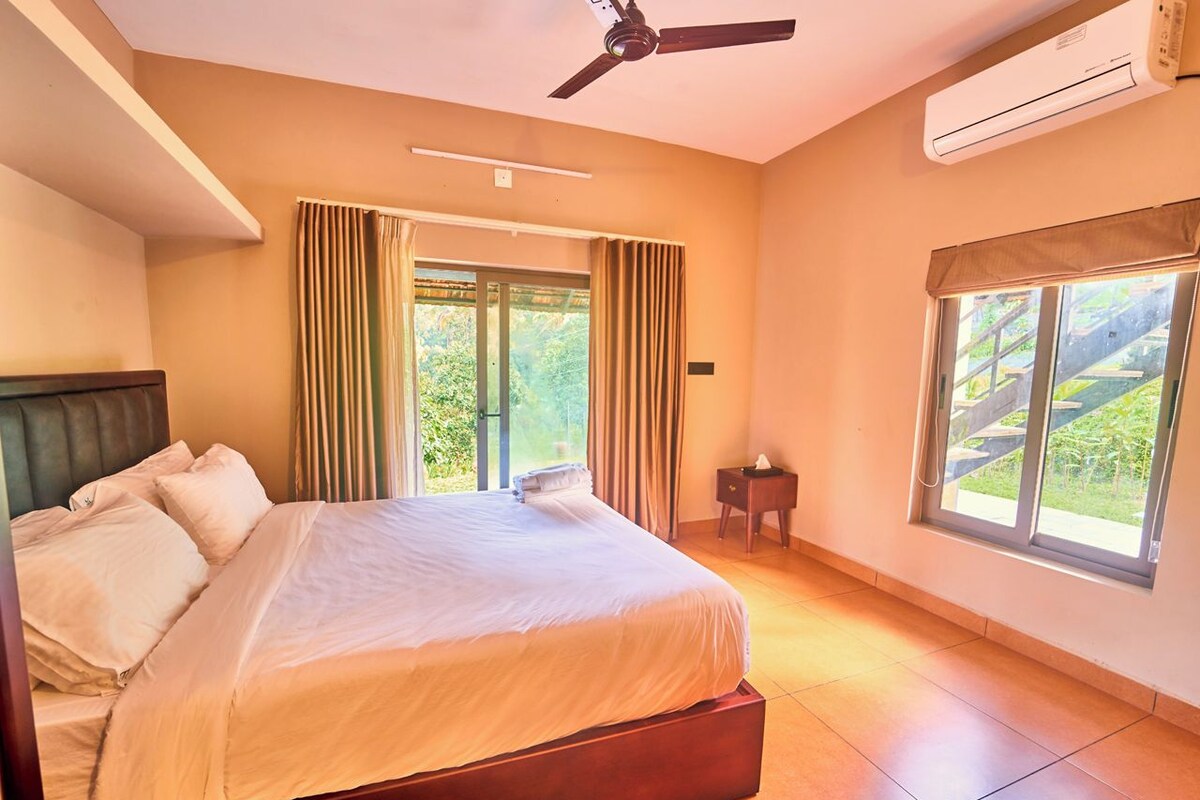 Quarto de hotel com uma cama grande, ventoinha no teto, aparelho de ar condicionado, uma mesinha e janela com vista para área verde.