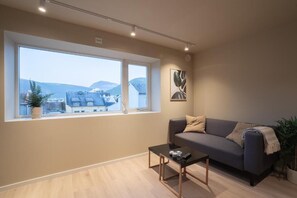 Uma sala de estar moderna com um sofá, uma mesa de centro e uma janela ampla com vista para edifícios e montanhas.