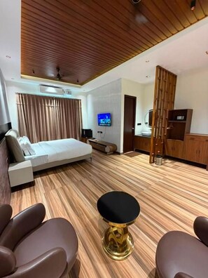 Quarto de hotel moderno com cama, televisão, piso de madeira e um espaço de estar com um banquinho.