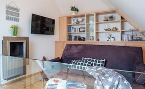 Uma sala de estar moderna com uma mesa de centro de vidro, um sofá roxo escuro e uma estante de madeira repleta de livros e objetos decorativos.