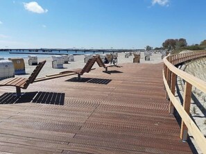Um calçadão de madeira com bancos e lixeiras, que leva a uma praia com um píer ao fundo.
