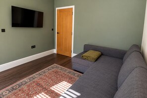 Uma sala de estar com sofá cinza, porta de madeira, TV de tela plana fixada na parede e tapete estampado no chão.