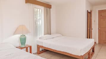 Quarto com duas camas de madeira, uma mesa de cabeceira com abajur, uma janela com cortinas e uma porta de madeira.