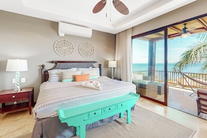 Quarto com cama, mesas de cabeceira, um banquinho, vista para o mar e ventilador de teto.