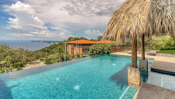Uma piscina infinita azul límpida com um guarda-sol de palha, vista para o mar e um edifício com telhado vermelho.