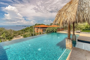 Uma piscina infinita azul límpida com um guarda-sol de palha, vista para o mar e um edifício com telhado vermelho.