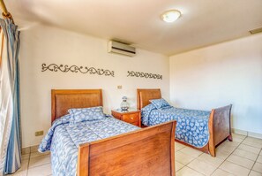 Quarto com duas camas de madeira, uma pequena mesa de cabeceira com um recipiente de vidro e um objeto decorativo na parede.