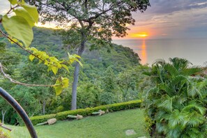 Paisagem exuberante com vista para o pôr do sol sobre a água, cercada por vegetação densa e árvores.