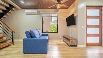 Uma sala de estar moderna com um sofá azul, piso de madeira, um ventilador de teto, uma TV na parede e uma escada.