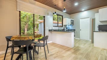 Uma cozinha moderna com uma ilha central, uma área de refeições com mesa e cadeiras de madeira, e vista para o exterior através de uma grande janela.