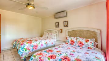 Quarto de hotel com duas camas, um ventilador de teto, ar condicionado e duas fotos emolduradas na parede.