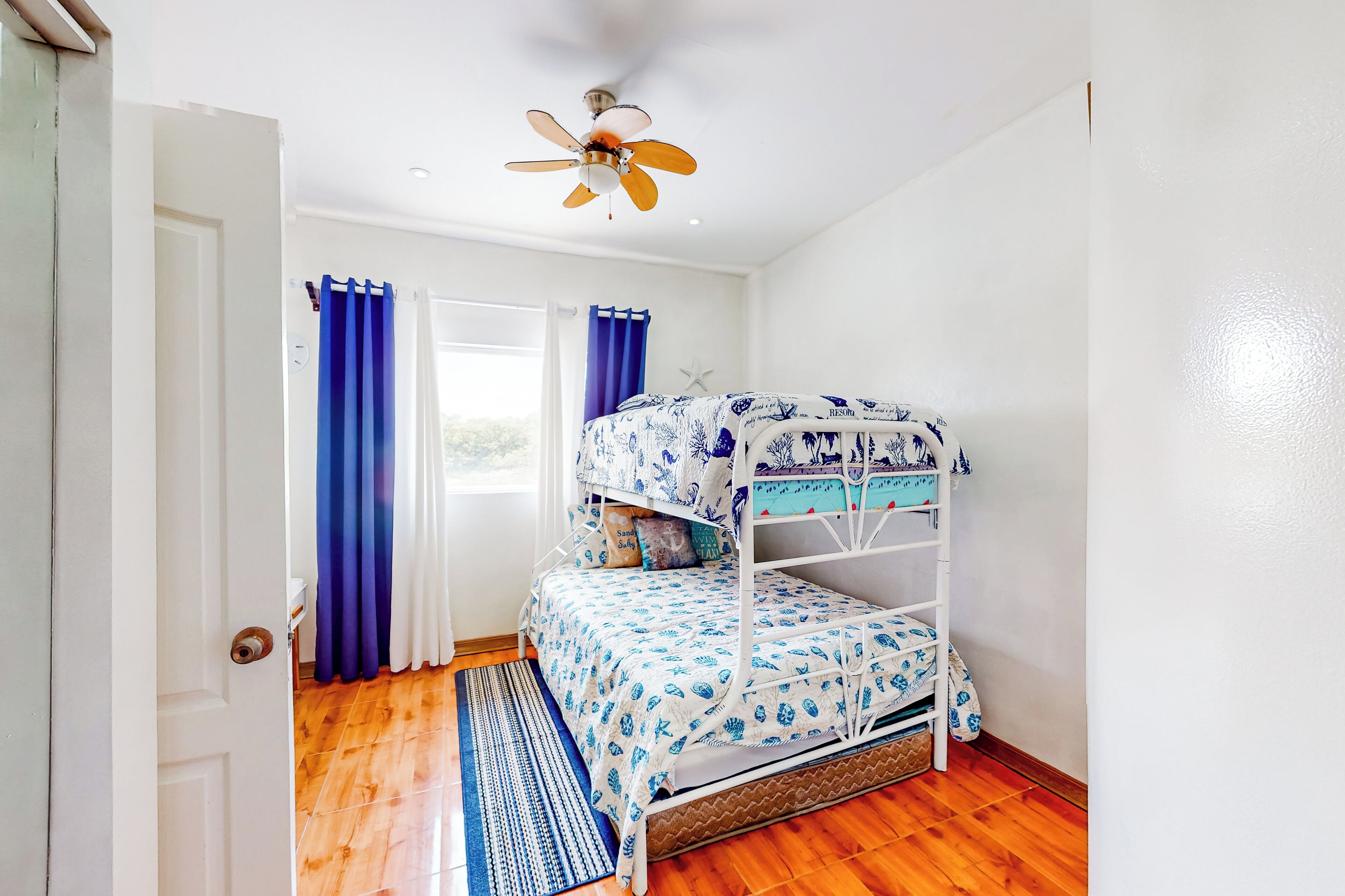 Uma cama de solteiro com edredom estampado, um ventilador de teto e uma janela com cortinas.