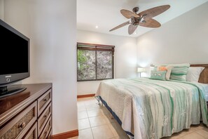 Quarto com cama, televisão sobre um armário de madeira, ventilador de teto e janela com persianas.