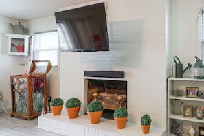 Uma sala de estar com lareira de tijolos brancos, uma televisão de tela plana montada acima e vários vasos com plantas dispostos na longa prateleira.