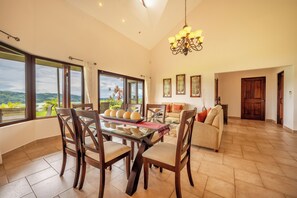 Ampla sala de estar com mesa de jantar, cadeiras e sofá. Grandes janelas oferecem vista para áreas verdes e colinas.
