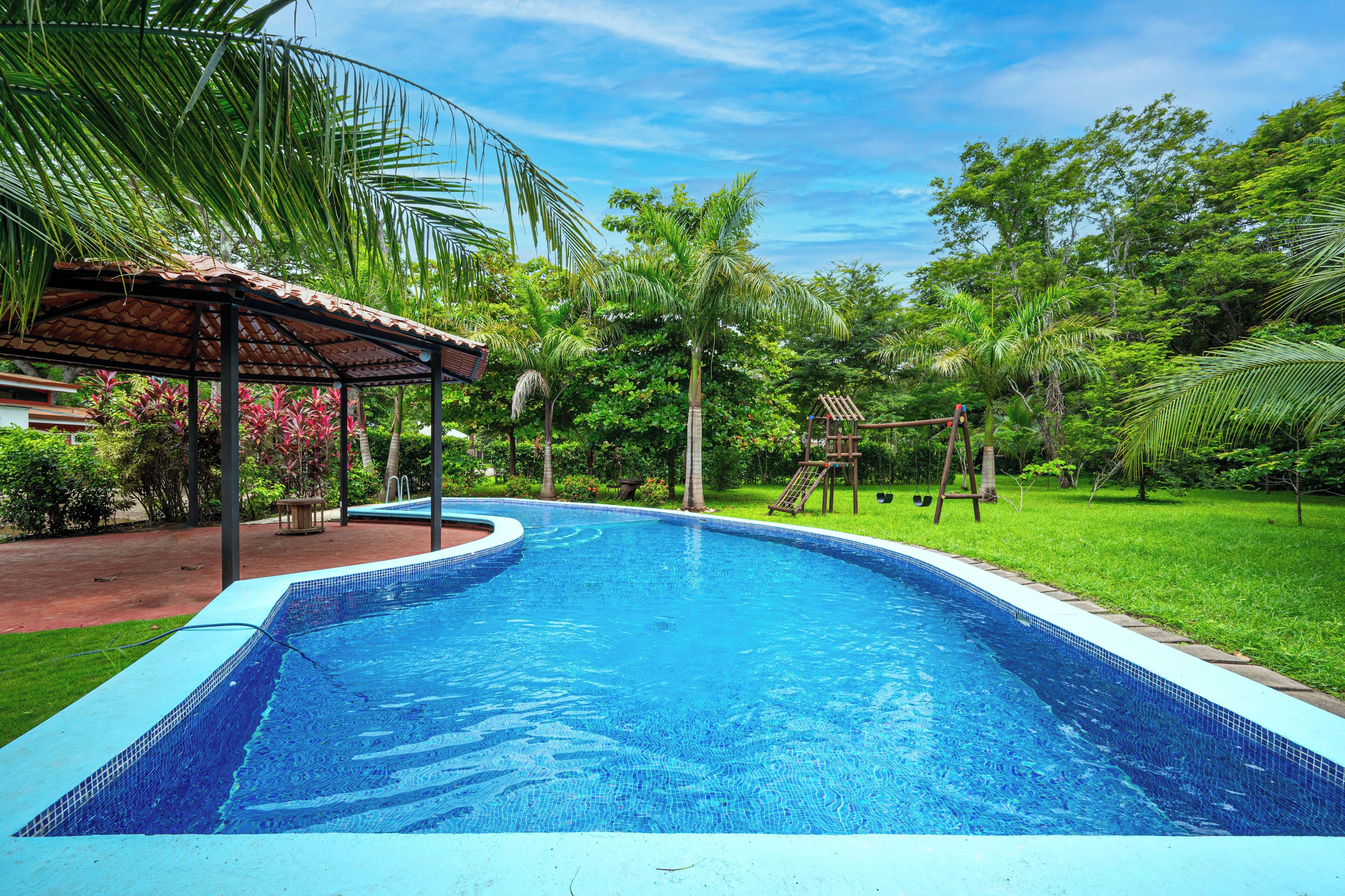 Piscina com borda em azulejo, rodeada por vegetação exuberante e uma área de estar coberta.
