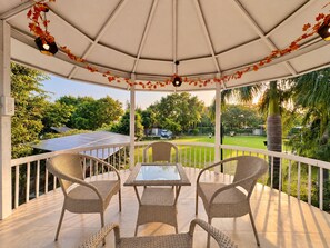 Área de estar coberta ao ar livre, com mobiliário de vime e mesa de vidro, com vista para um jardim e painéis solares.