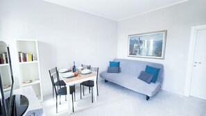 Um espaço de estar moderno com área de jantar, sofá cinza com almofadas azuis, estante de livros e uma pintura emoldurada de paisagem urbana na parede.
