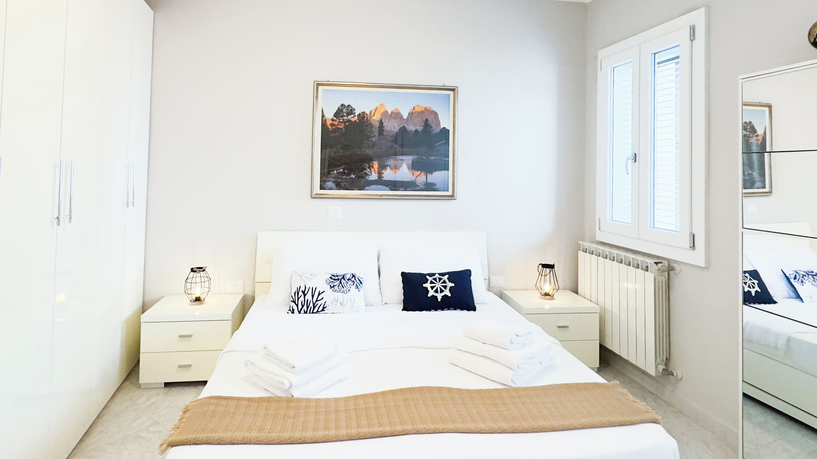 Quarto de hotel com cama, duas mesinhas de cabeceira, um radiador, uma janela com persianas e um quadro na parede.