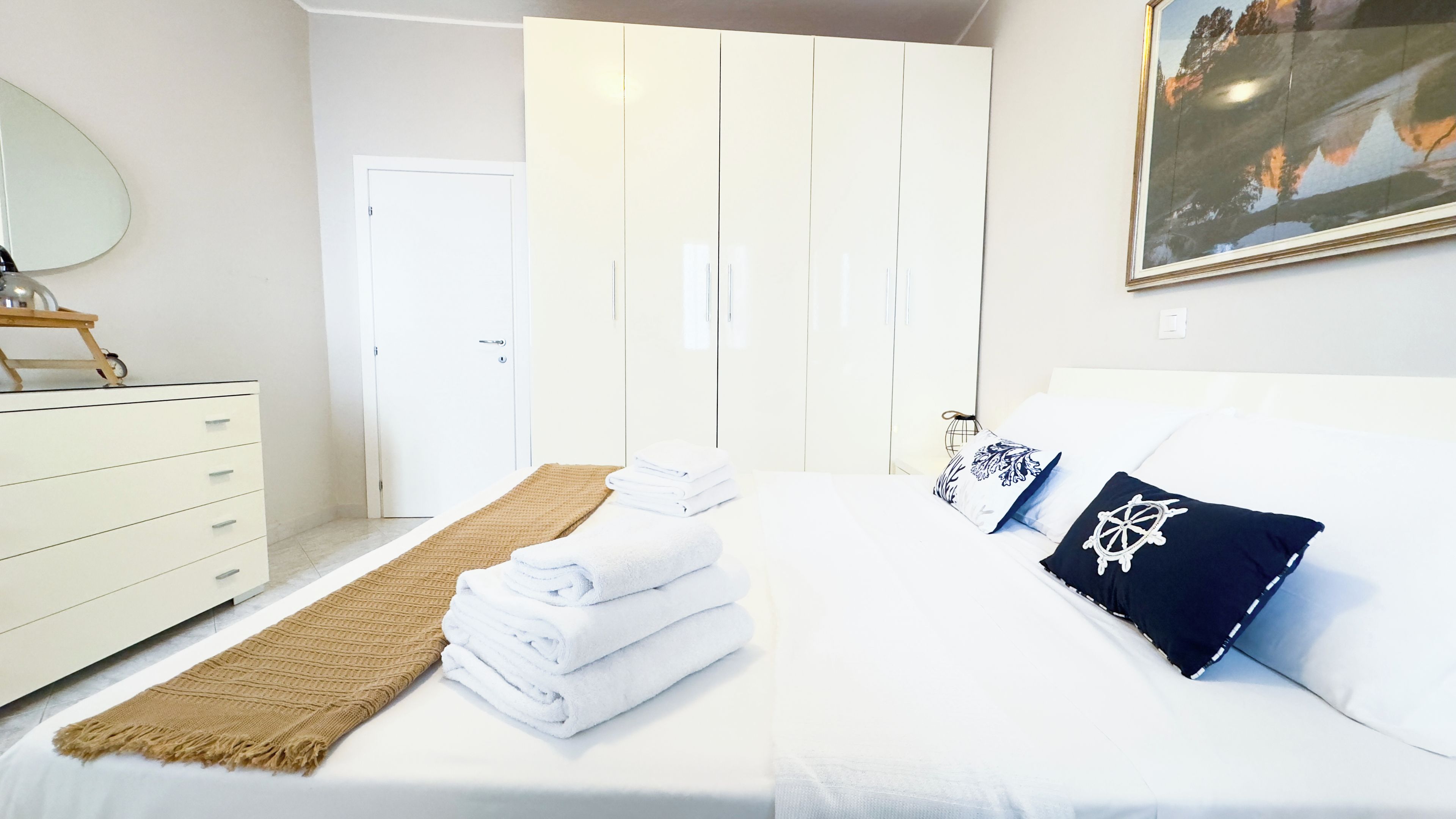 Quarto de hotel com cama, penteadeira branca, guarda-roupa e um quadro na parede.