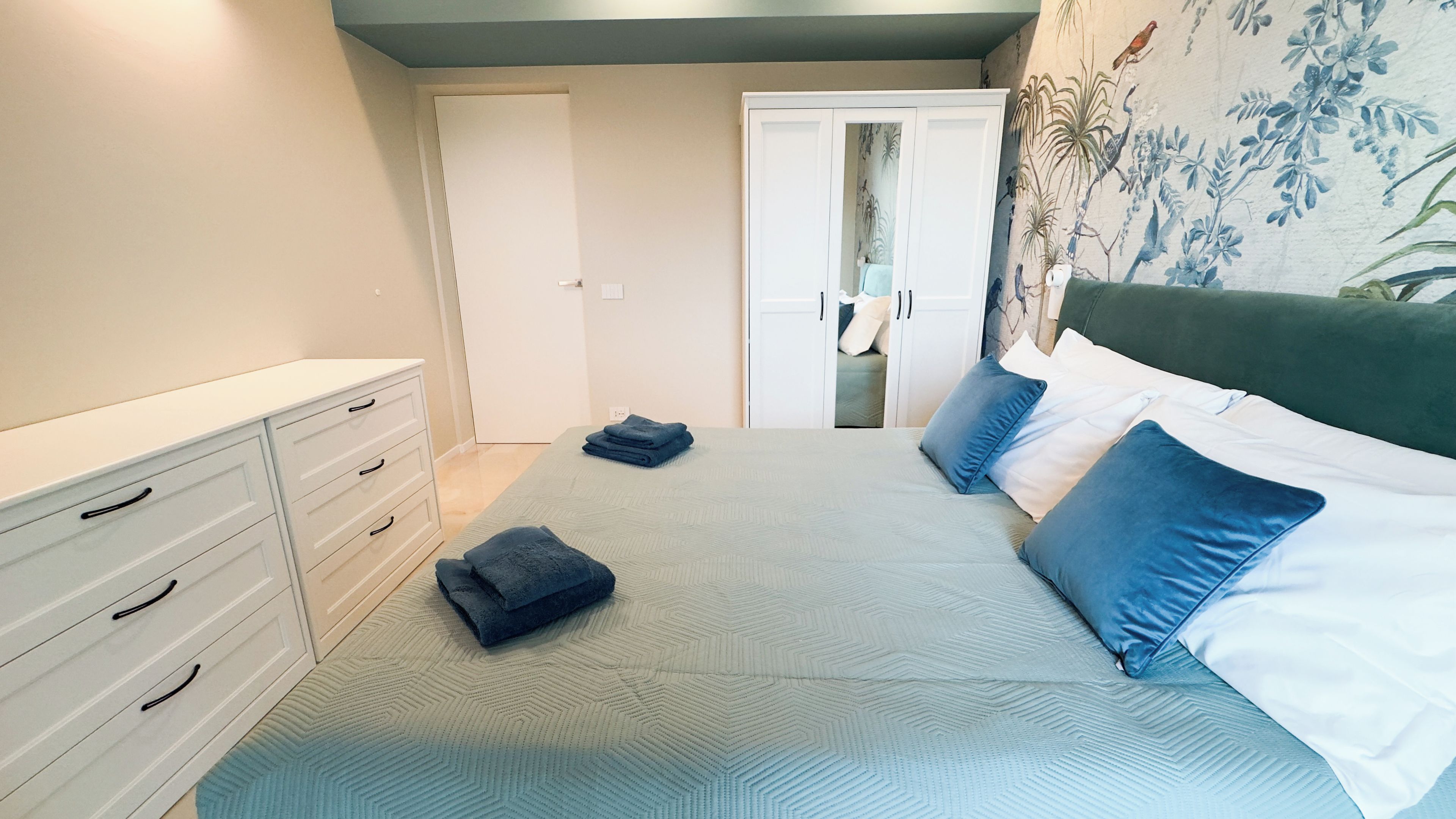 Quarto com cama, penteadeira e porta que dá para outro cômodo.