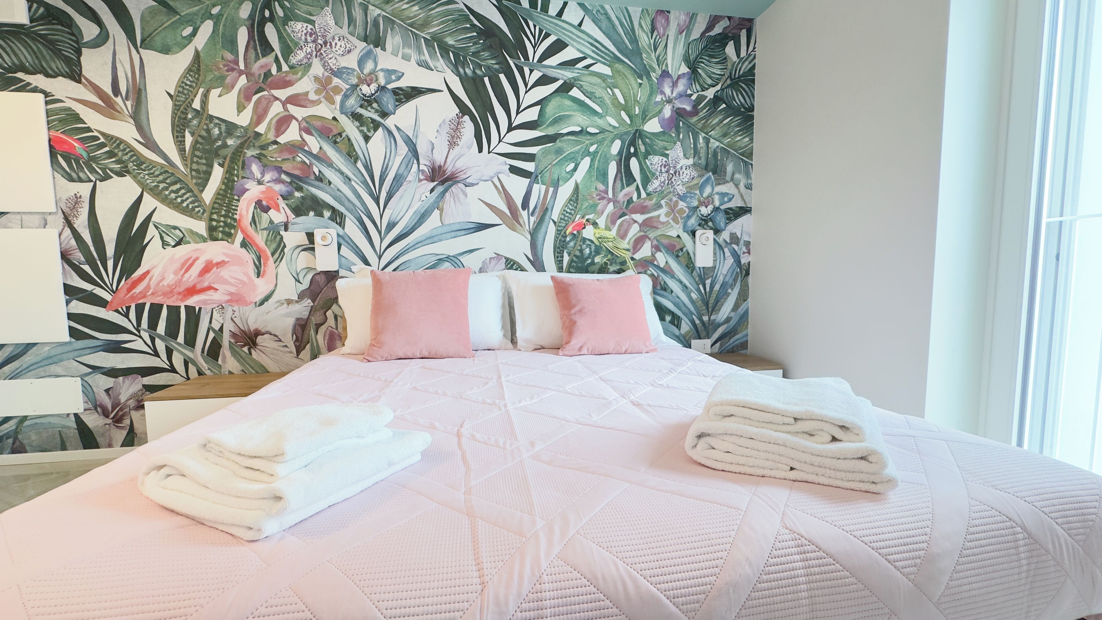 Quarto de hotel com cama, almofadas e papel de parede tropical.