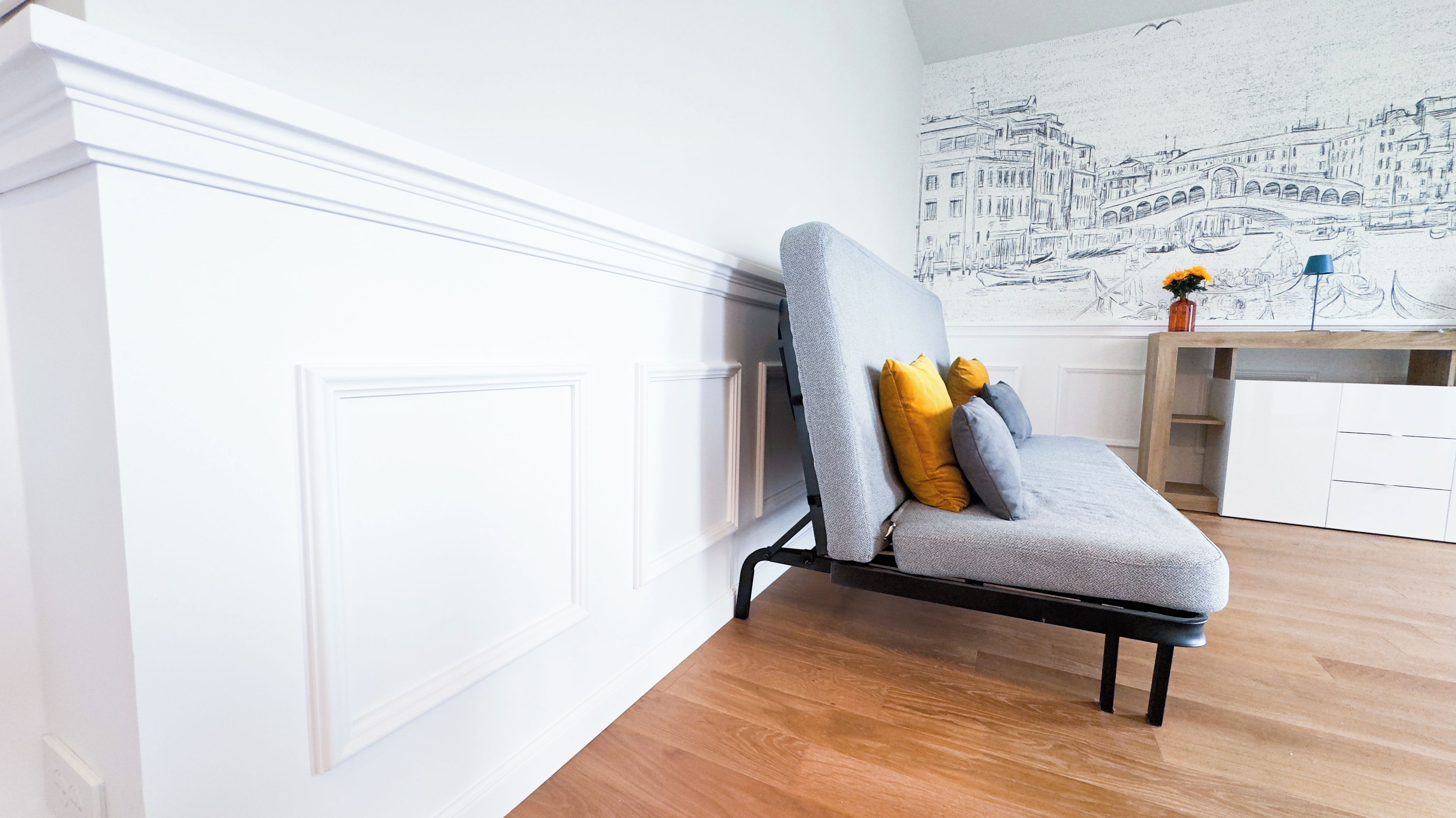 Quarto moderno com sofá cinzento, parede branca, piso de madeira e um mural com vista para a cidade.
