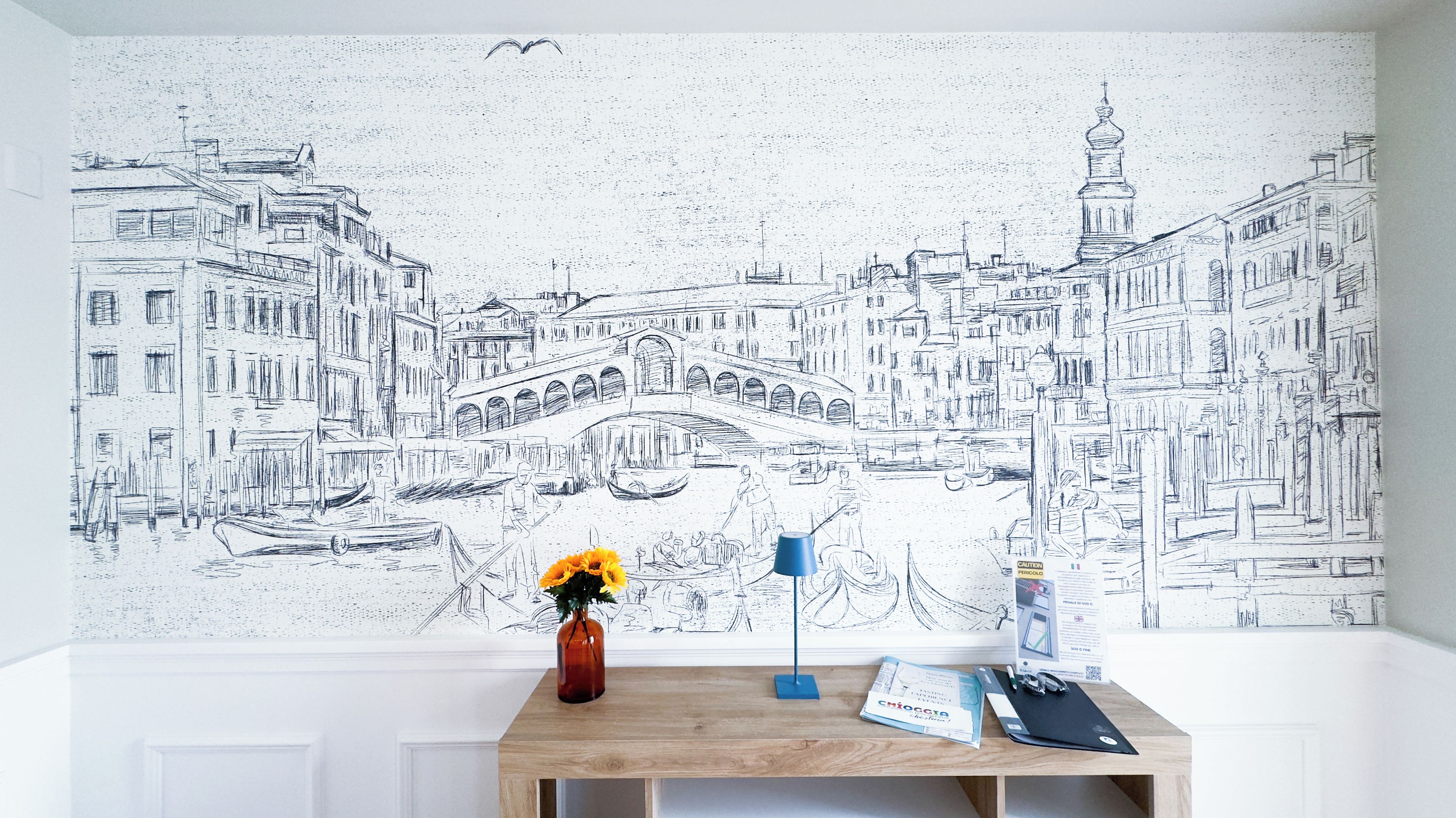 Um mural na parede retrata uma paisagem urbana com uma ponte, uma escrivaninha com um vaso de flores, uma luminária e alguns papéis.