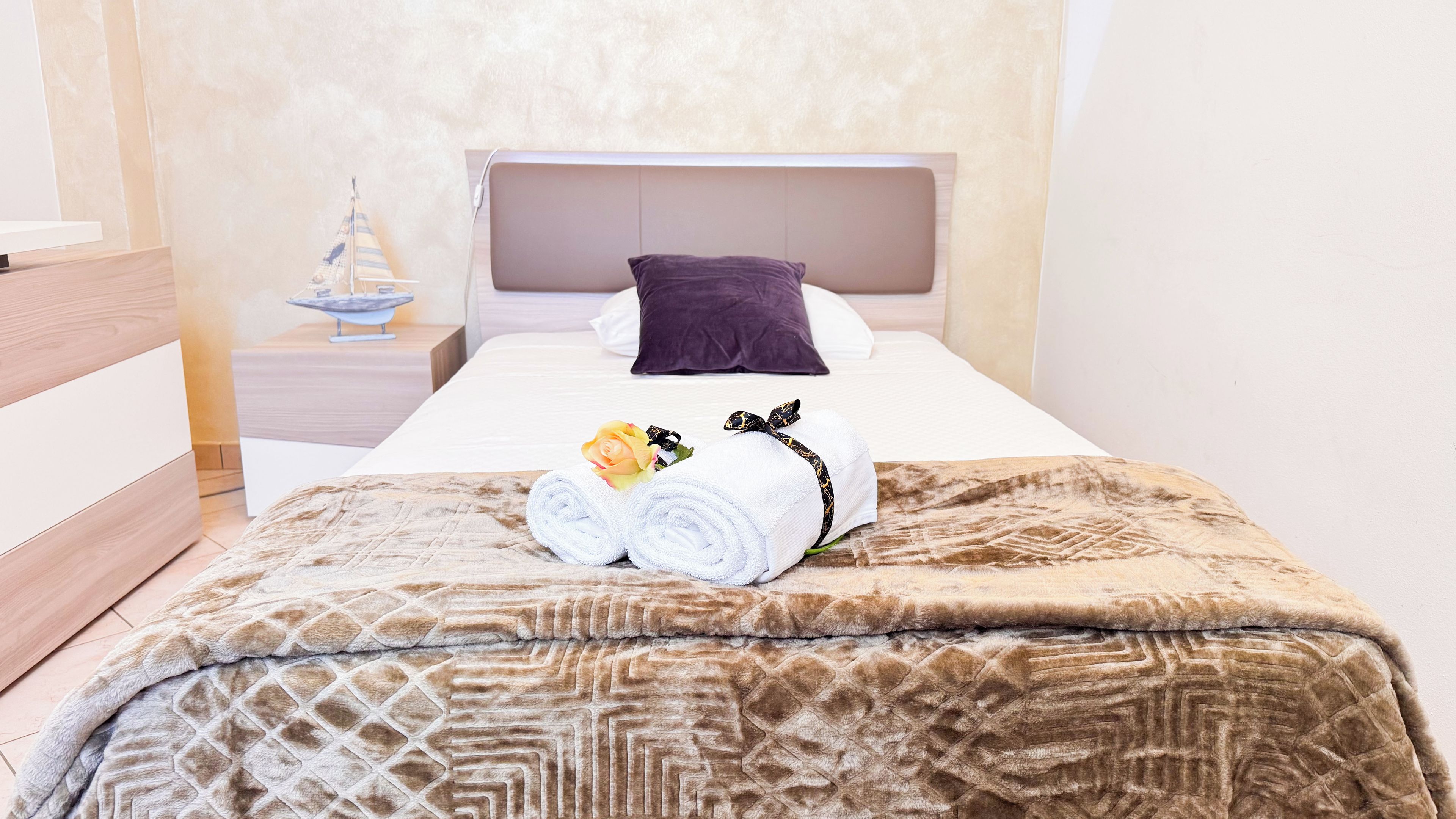 Uma cama bem arrumada com um cobertor marrom estampado, uma toalha branca enrolada, uma rosa e um laço preto e dourado.