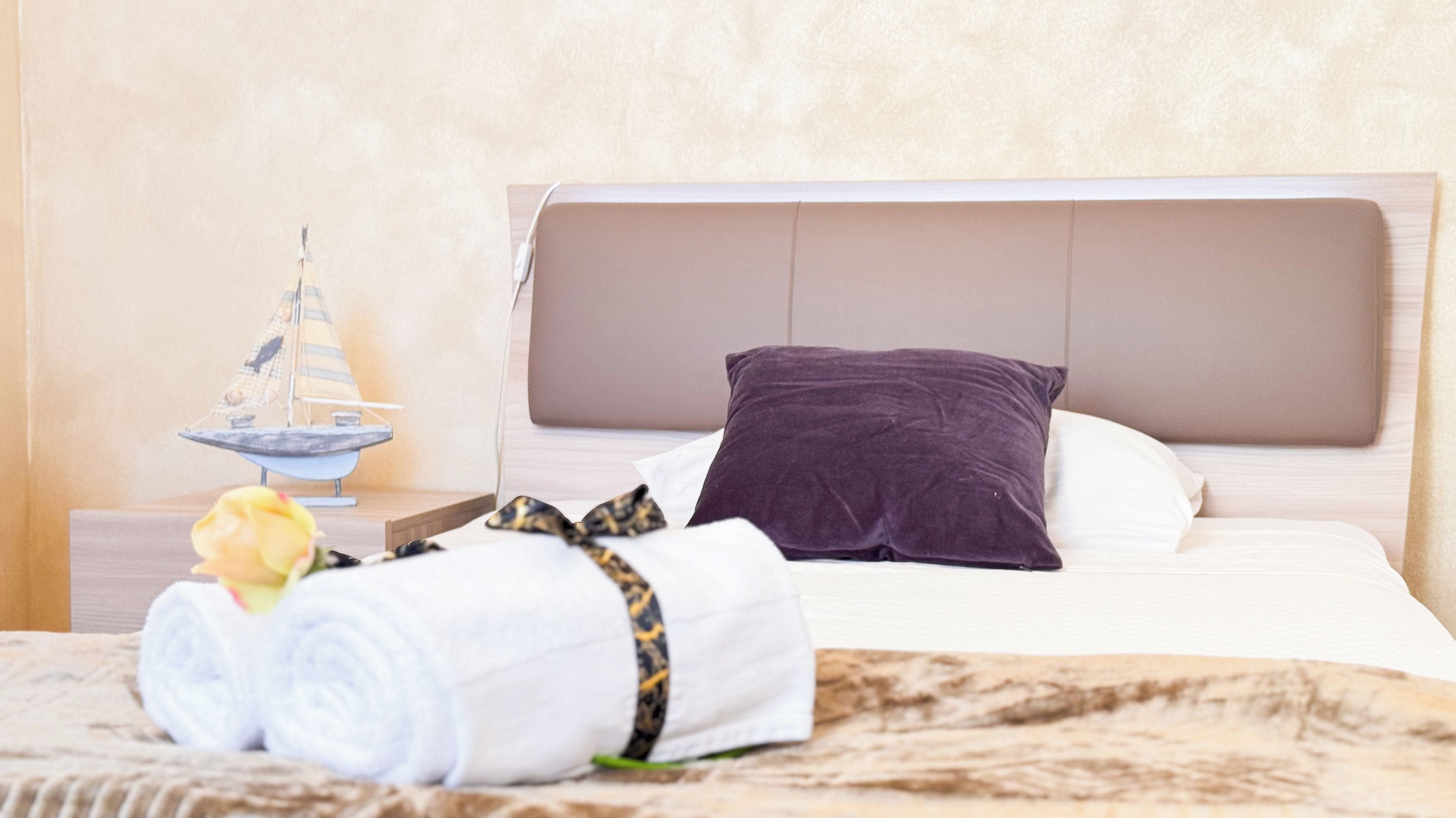 Cama bem arrumada com cabeceira, toalha enrolada, travesseiro roxo e um pequeno barco à vela decorativo sobre a mesa de cabeceira.