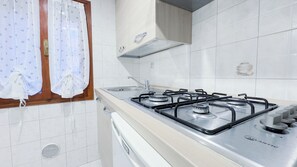 Cozinha com paredes em azulejo branco, fogão a gás de aço inoxidável, pia e janela com cortina.
