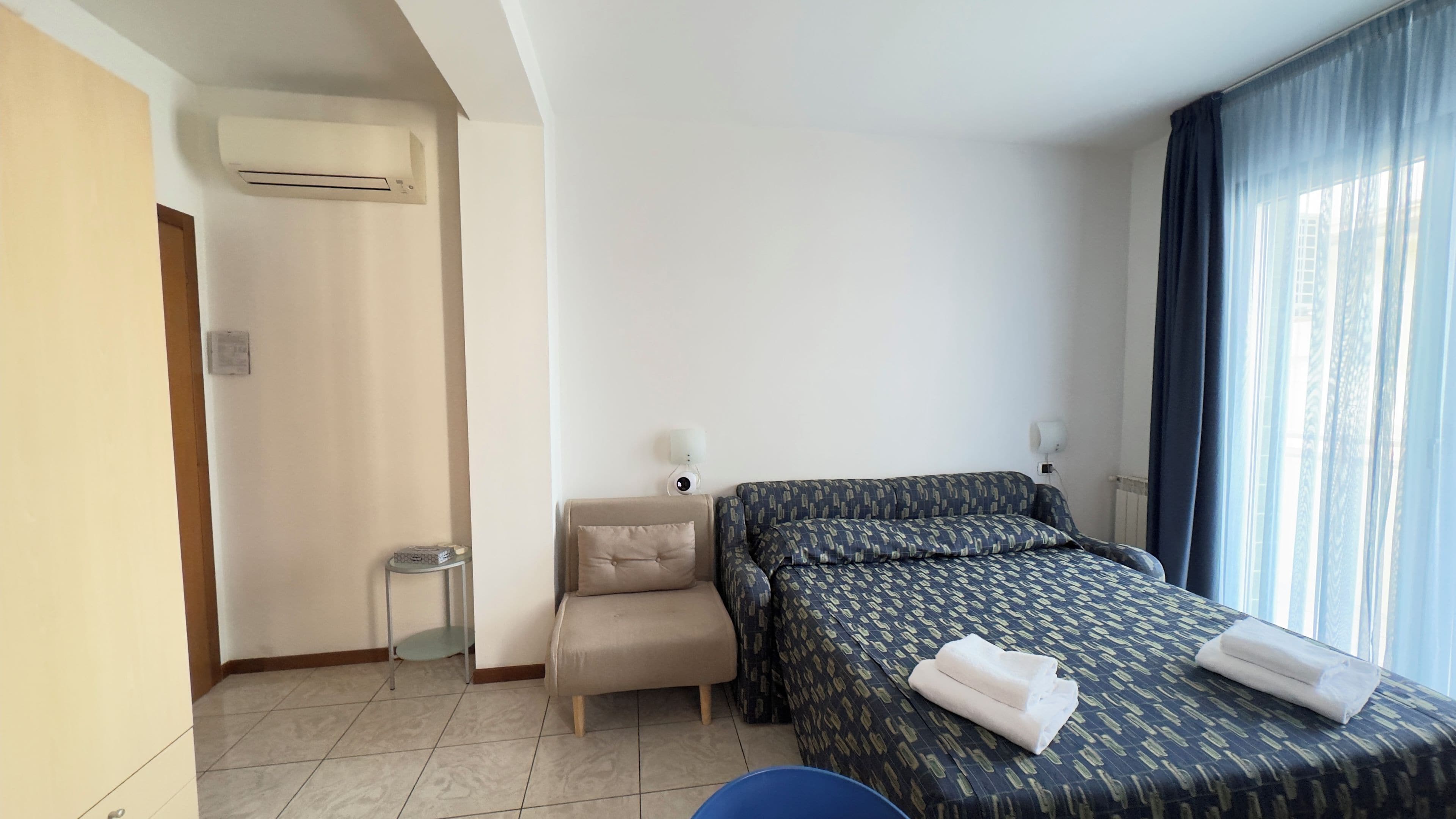 Quarto de hotel com cama, cadeira, ar condicionado, mesa pequena e janela com cortinas.