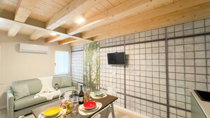 Quarto com teto de madeira, TV fixada em parede revestida de azulejos, mesa de jantar posta com pratos, copos e uma garrafa, e um sofá com almofada.