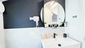 Casa de banho moderna com lavatório branco, secador de cabelo fixado na parede e espelho refletindo uma planta em vaso e uma janela.