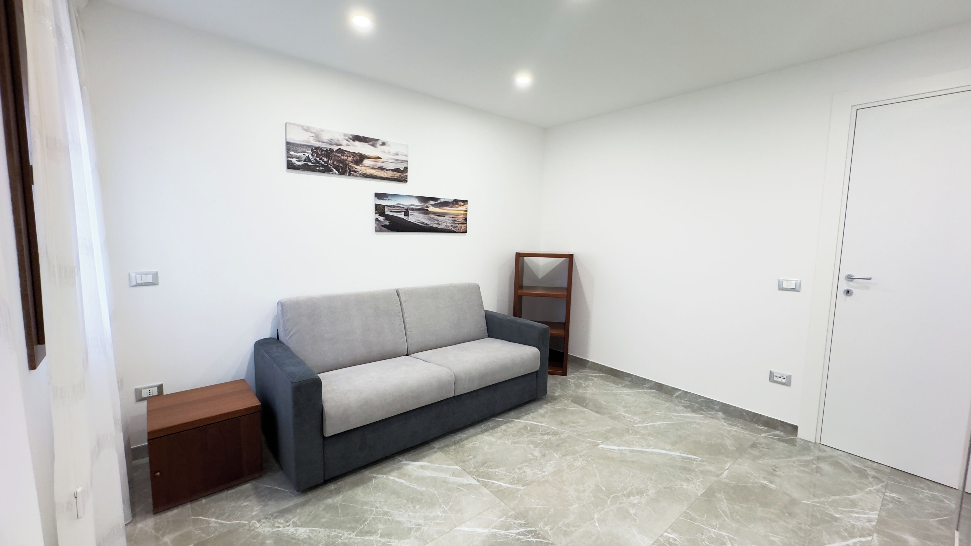 Uma sala de estar moderna com um sofá cinzento, uma mesinha de madeira, piso de mármore e uma porta.