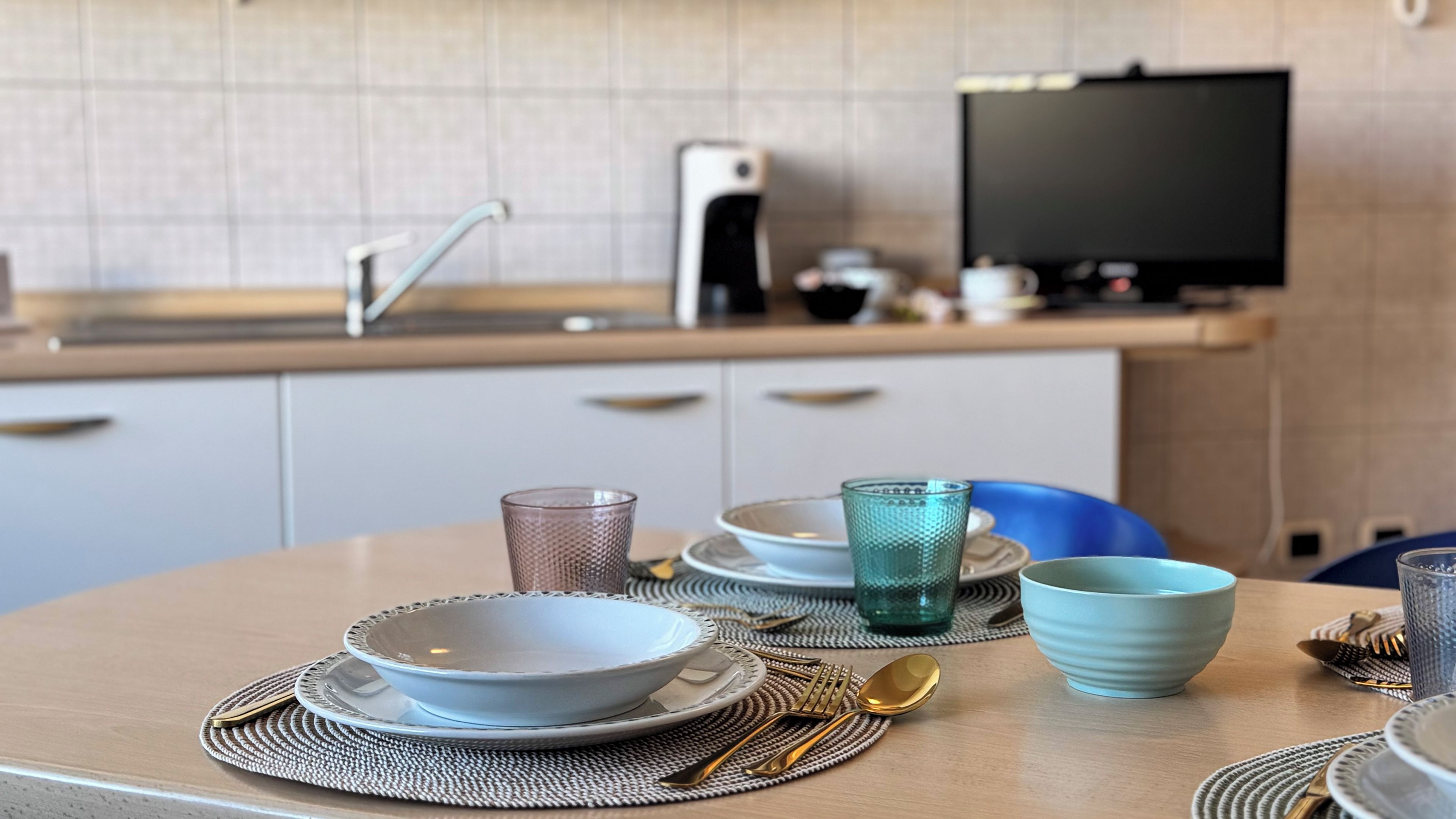 Uma cozinha moderna com uma mesa de jantar posta para dois, apresentando uma TV de tela plana, uma cafeteira e um revestimento de azulejos.